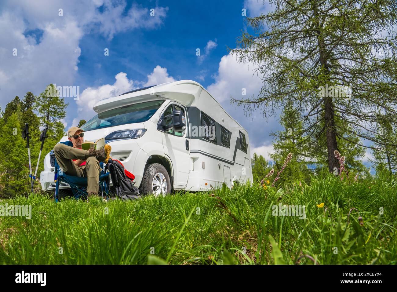 A man relaxes in a camping chair by a white campervan, enjoying a book ...