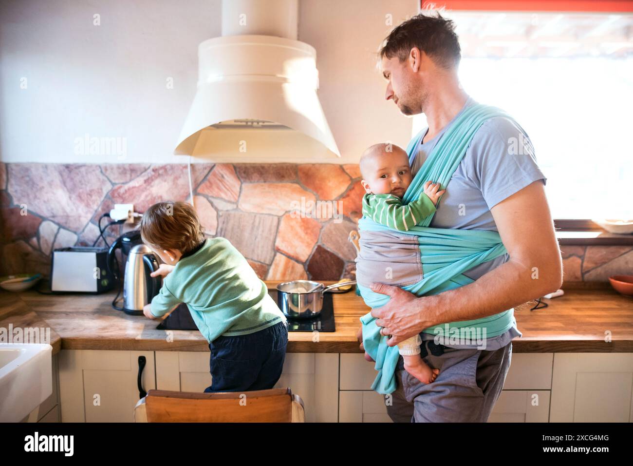 Father holding small baby in baby sling. Older brother stirring ...