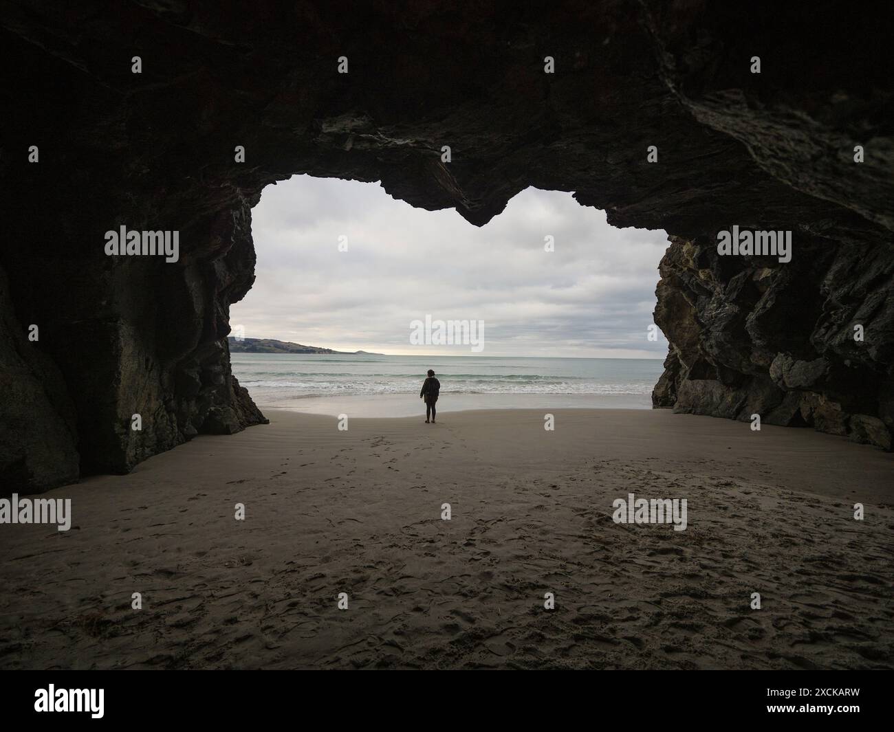 A person standing in large natural rock sea cave arch opening with sand ...