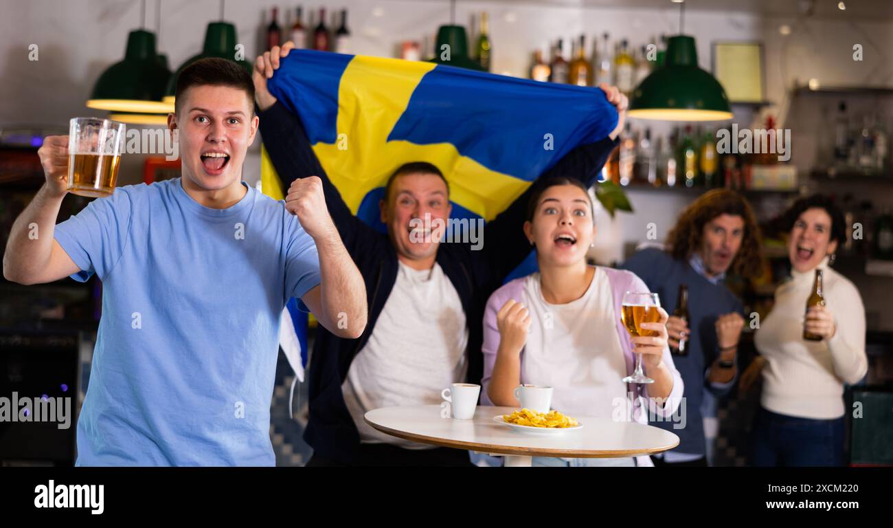 Company of excited young adult sports fans waving flag of Sweden and ...