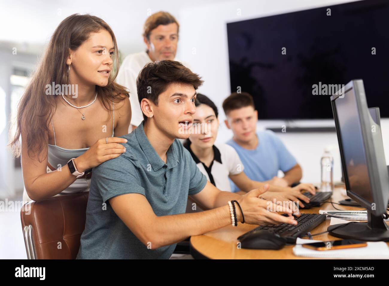 Girl stands behind sitting students, looks at monitor and helps in ...
