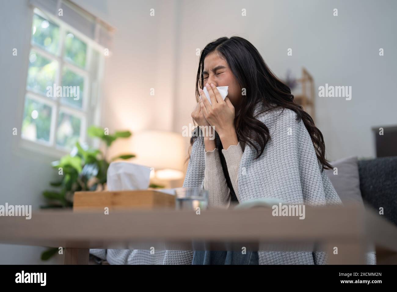 Young woman with cold and runny nose sneezing into tissue while wrapped ...