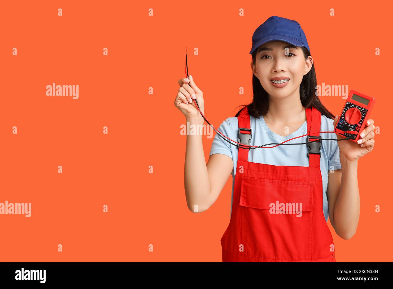 Young female electrician with multimeter and wires on orange background ...