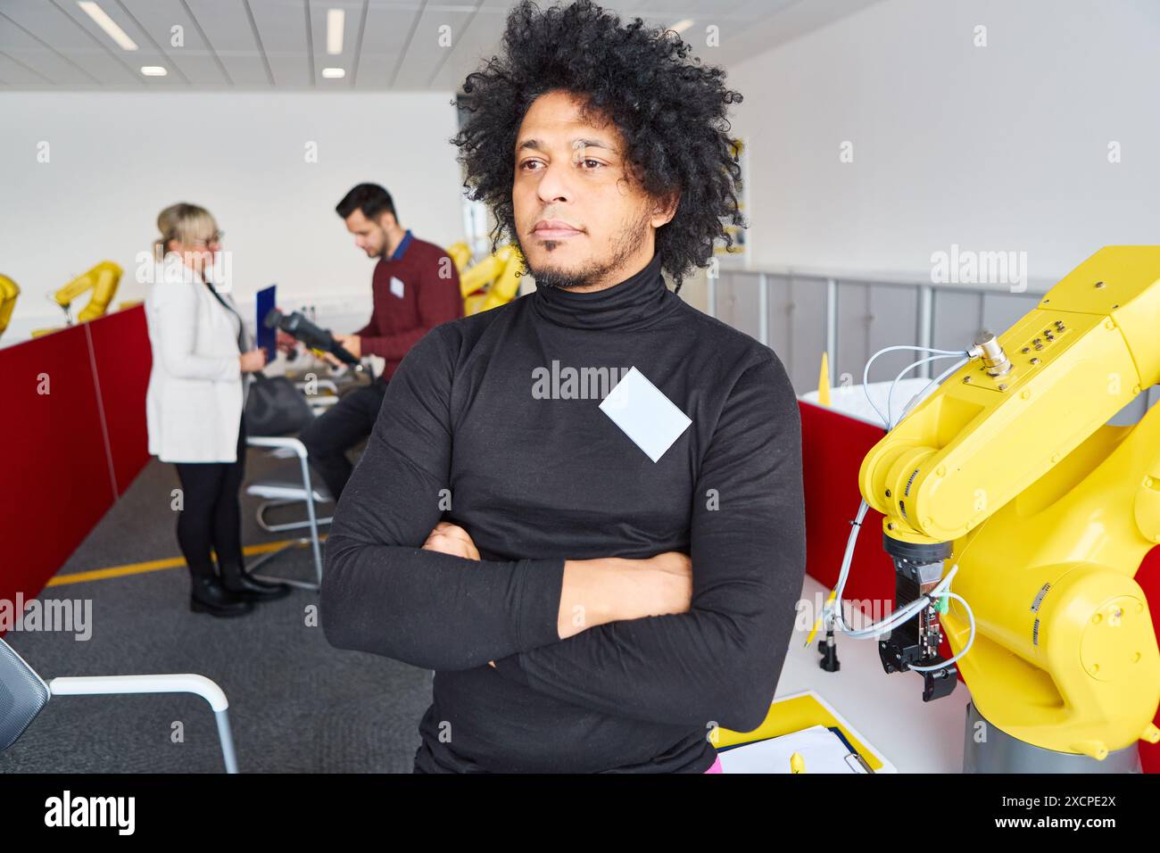 Engineer stands confidently next to a robotic arm in a laboratory. Colleagues work in the ...
