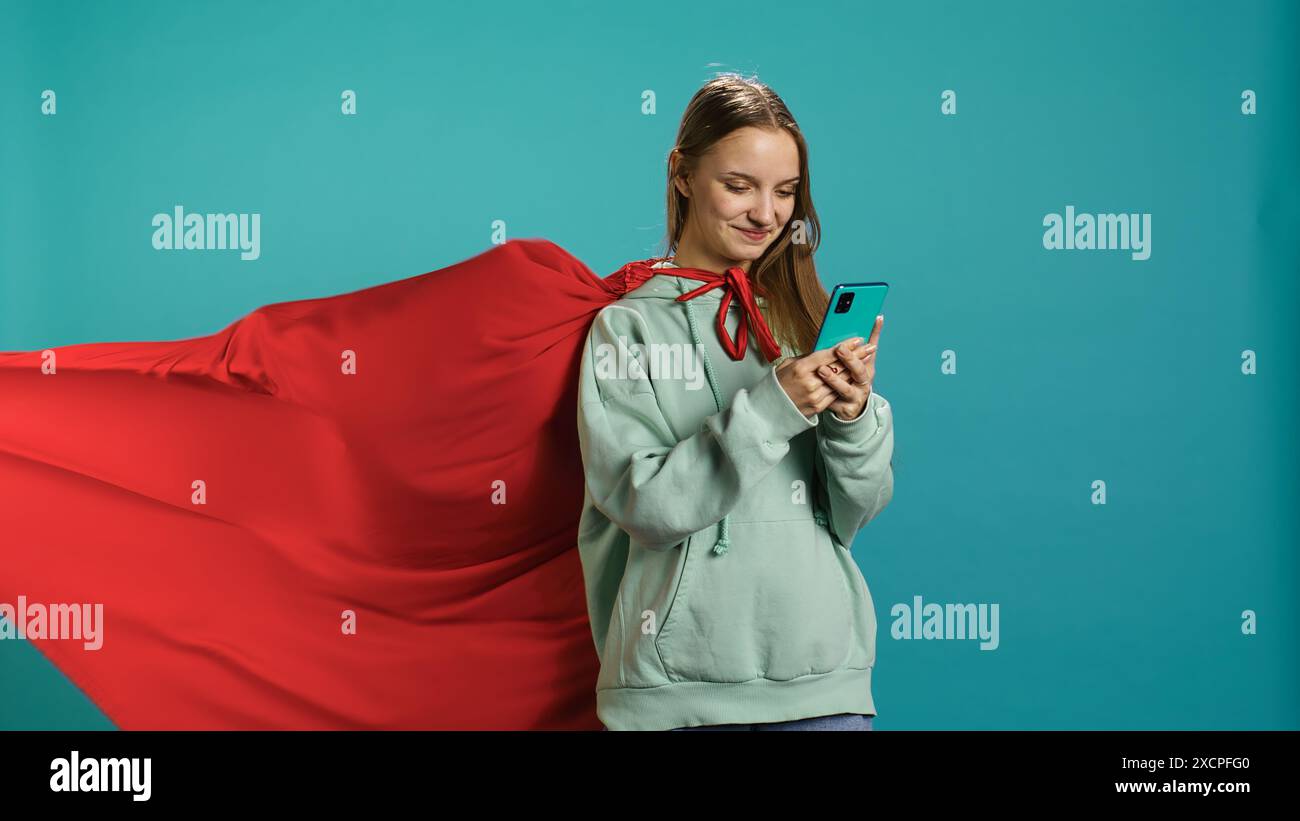 Young girl wearing superhero costume texting friends using smartphone, studio backdrop. Woman ...