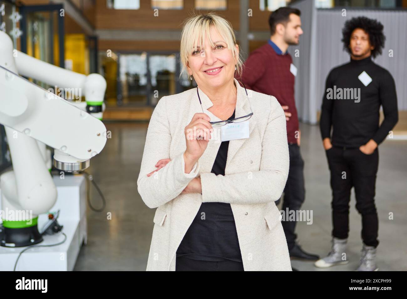 Confident professional woman posing beside robotic arm, with colleagues ...
