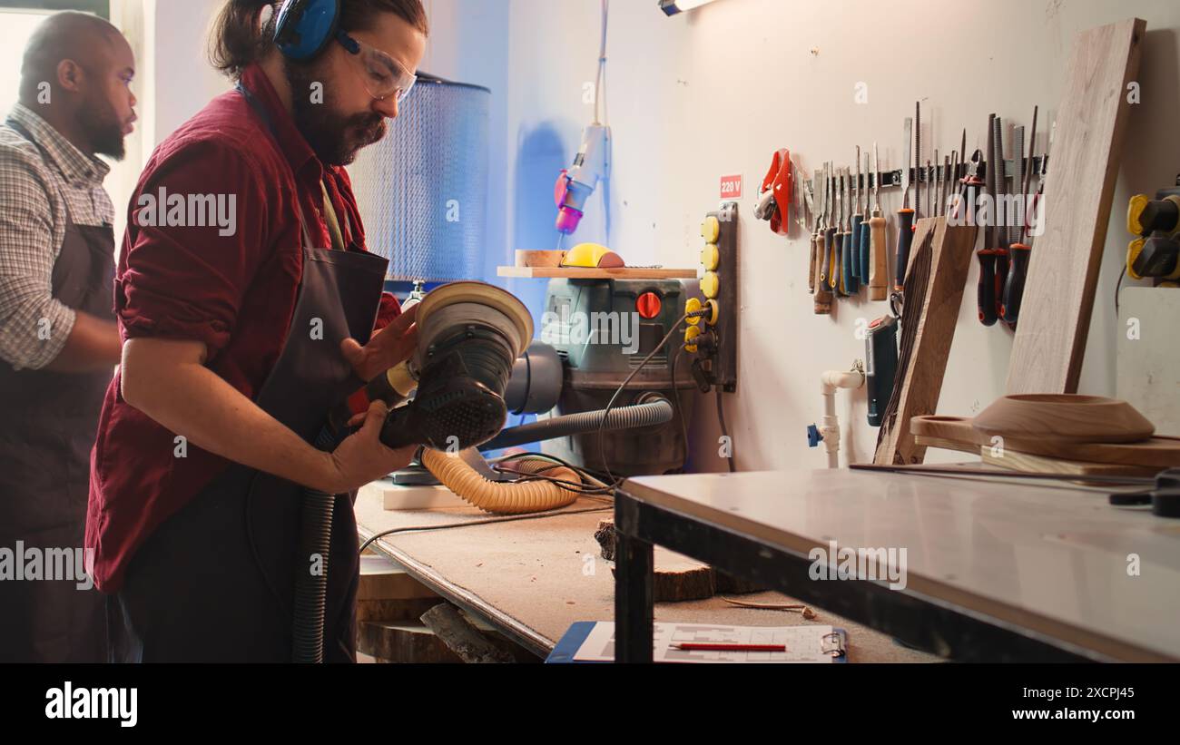 Carpenter at work consulting blueprint, using orbital sander on lumber ...