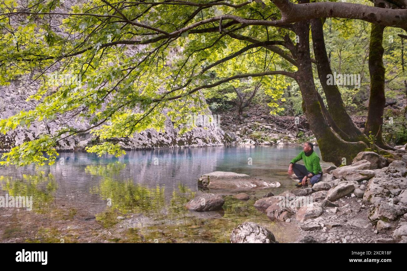 A man sits by the edge of the crystal-clear Voidomatis River, surrounded by lush greenery and ...
