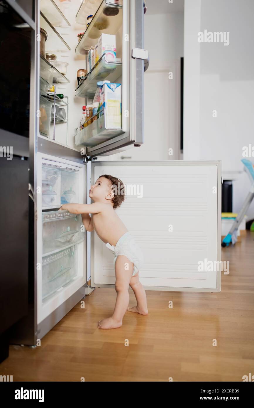 A young child, wearing a diaper, stands on tiptoes to reach inside an ...
