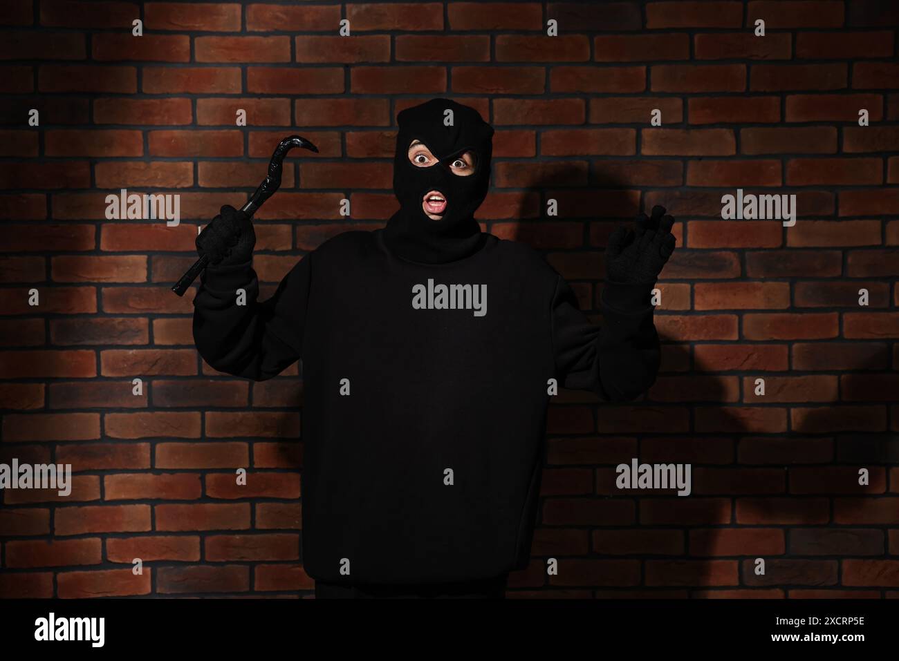Emotional thief in balaclava raising hands with crowbar against red ...