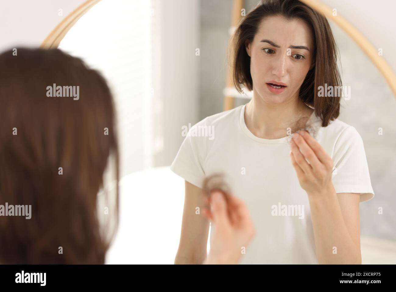 Stressed woman holding clump of lost hair near mirror indoors. Alopecia ...
