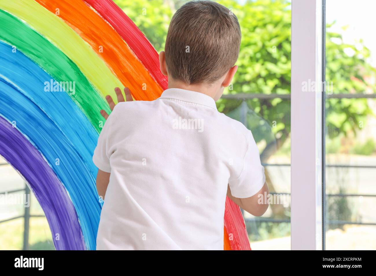Little boy touching picture of rainbow on window indoors, back view ...
