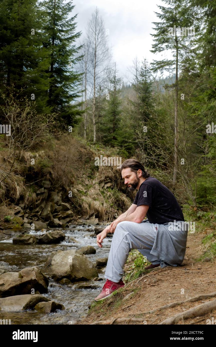 caucasian man sits in the forest near a mountain river and resting ...