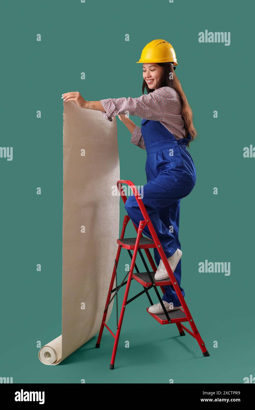 Female Asian decorator with wallpaper on stepladder against green ...