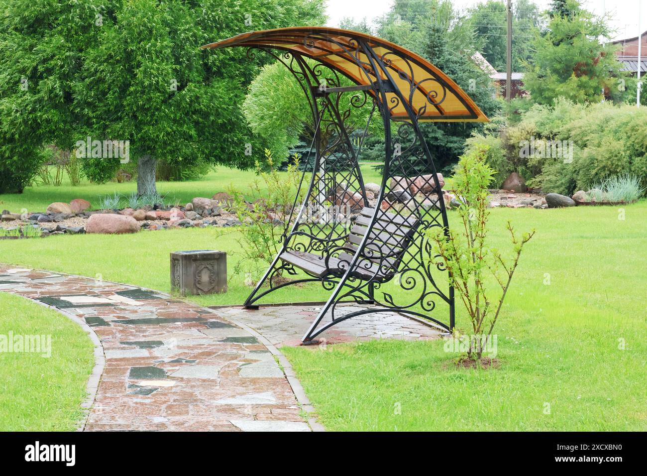 Wrought iron swing with canopy in park near stone path. Recreation in ...