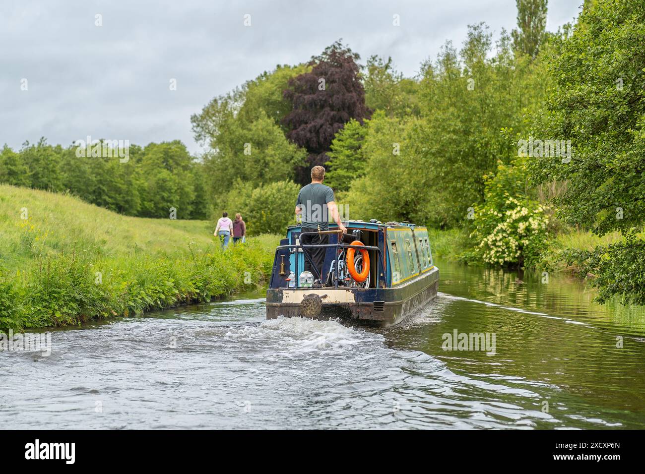 Rear view of a man steering a moving canal boat along a rural UK canal ...