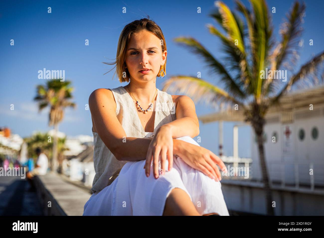 A serious young woman is sitting on a ledge by the ocean, wearing a ...