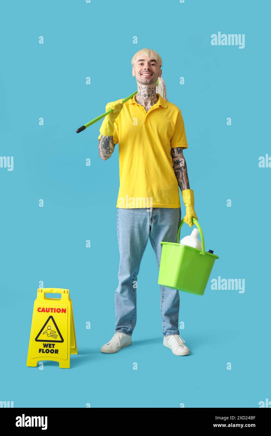 Young tattooed man with cleaning supplies and caution sign on blue ...