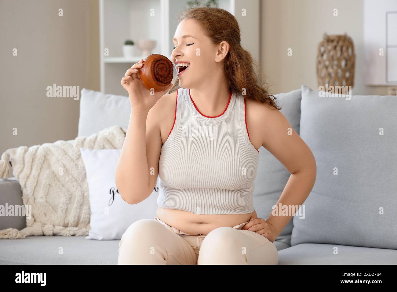 Young woman in tight pants eating bun on sofa at home. Weight gain ...