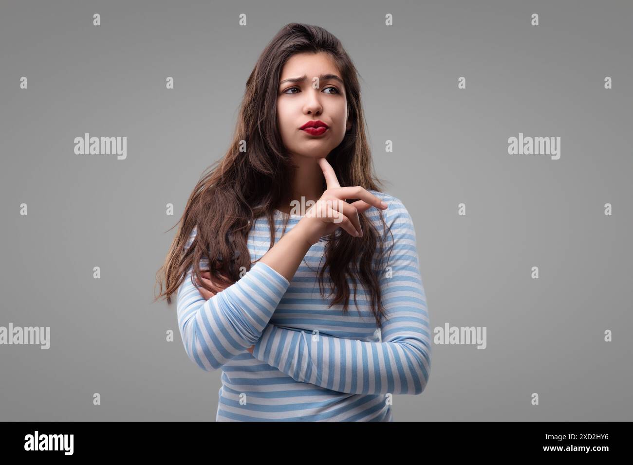 Young woman looks thoughtful, holding her chin, contemplating a ...