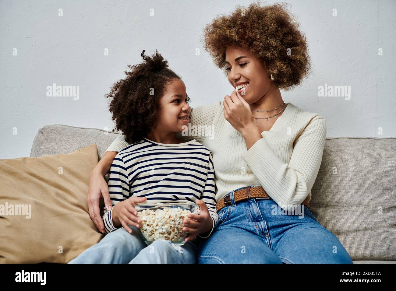 Happy African American mother and daughter sit on a couch, enjoying a snack of popcorn together ...