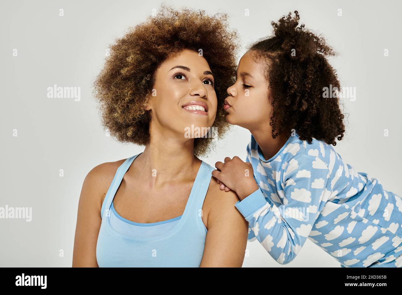 Happy African American mother and daughter wearing pajamas share a tender kiss in front of a ...
