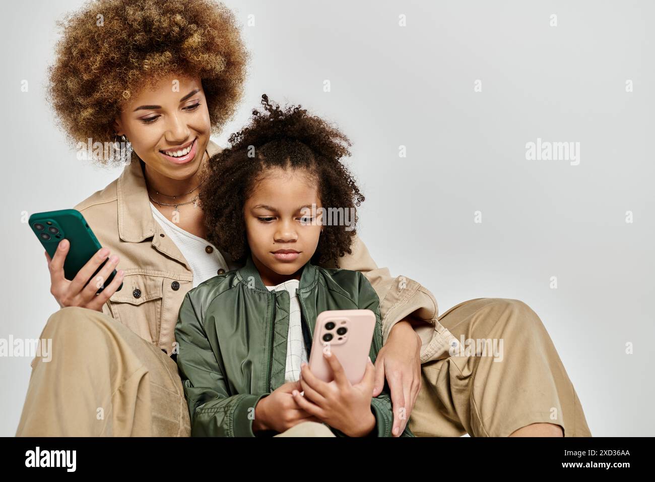 Curly African American mother and daughter in stylish clothes using cell phones while seated on ...