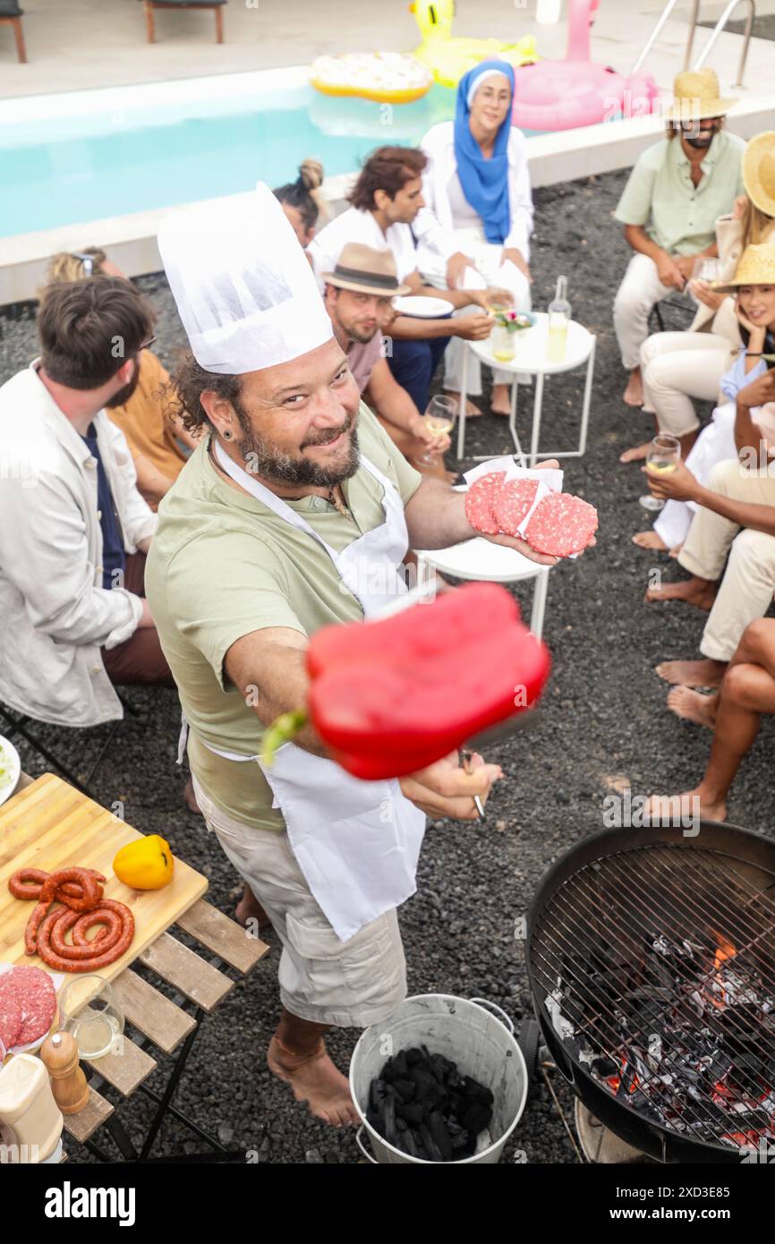 A multiethnic gathering of friends around a barbecue grill by the pool, with a man in a chef hat ...