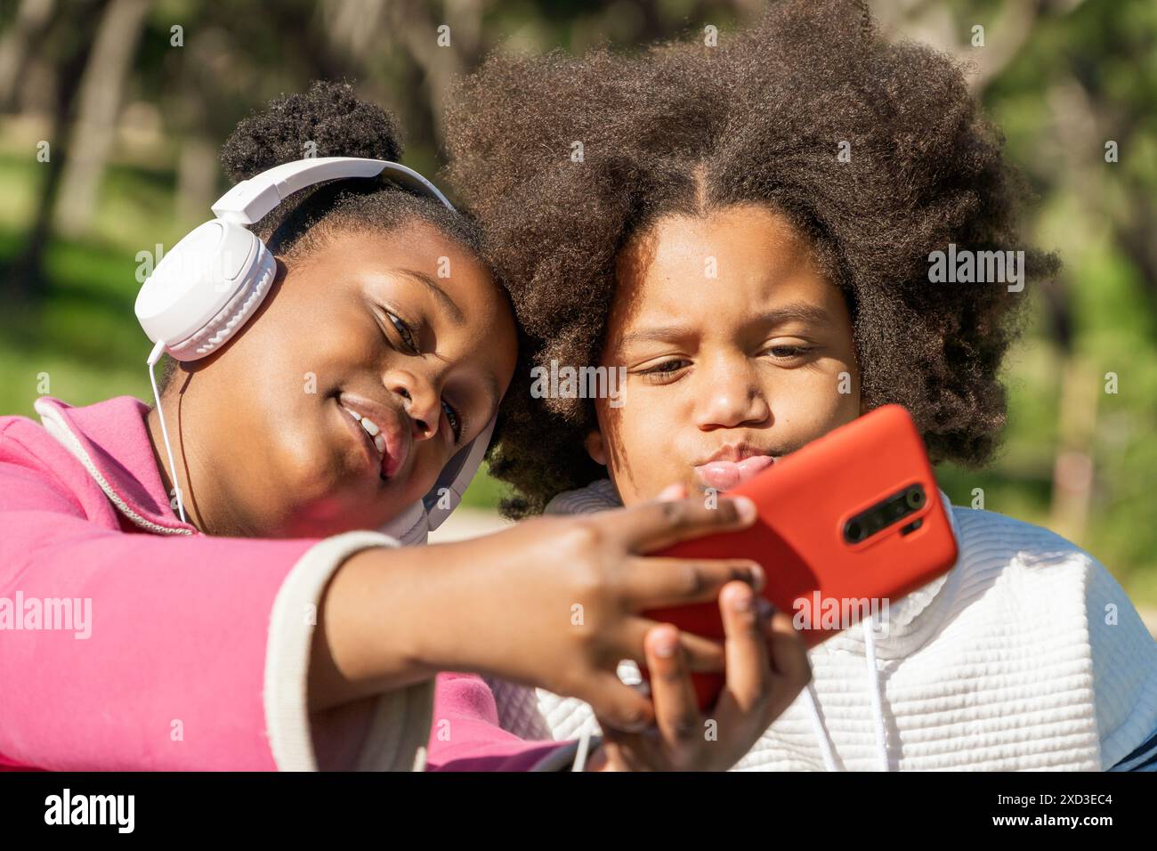 Two joyful girls taking selfies in a sunny park One giggles while the ...