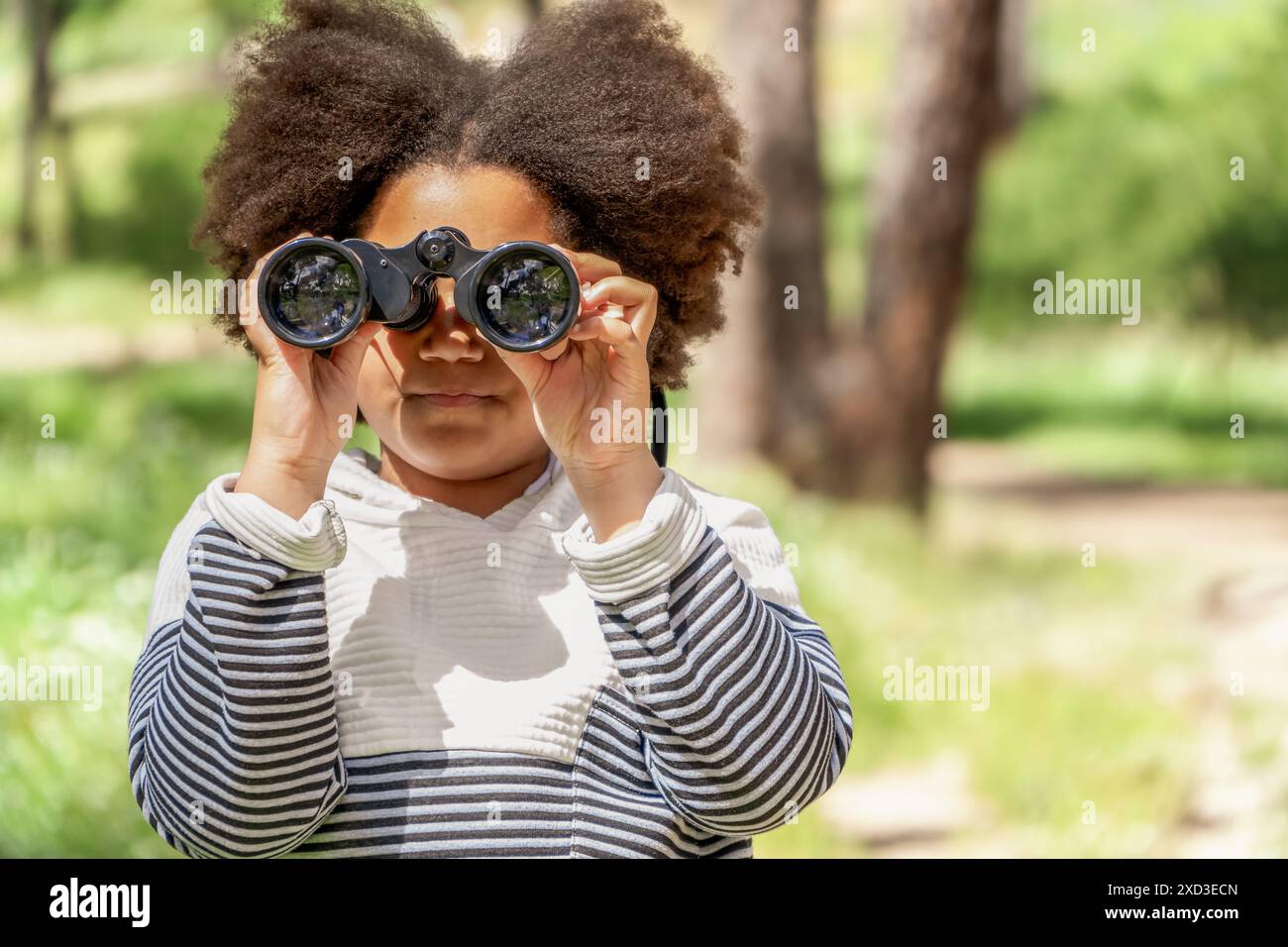 A young African American girl uses binoculars to explore her surroundings in a lush park ...