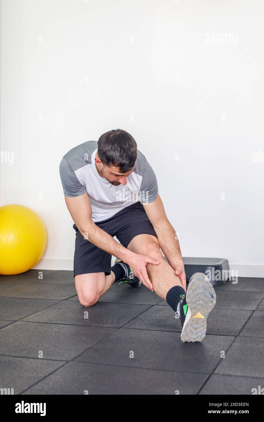 A focused man in a gym stretches his leg, grasping his calf, with a ...