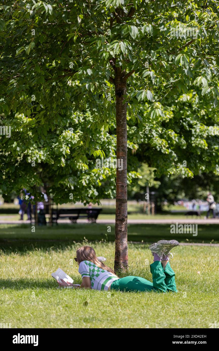 A young woman sunbathes whilst reading her book sheltering under a tree ...