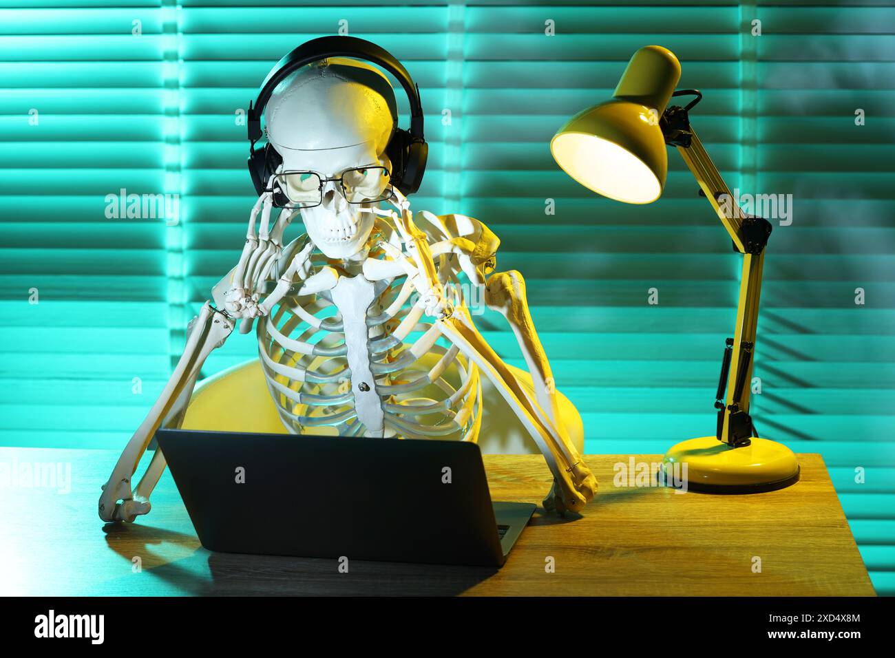 Waiting concept. Human skeleton sitting at wooden table with laptop and ...