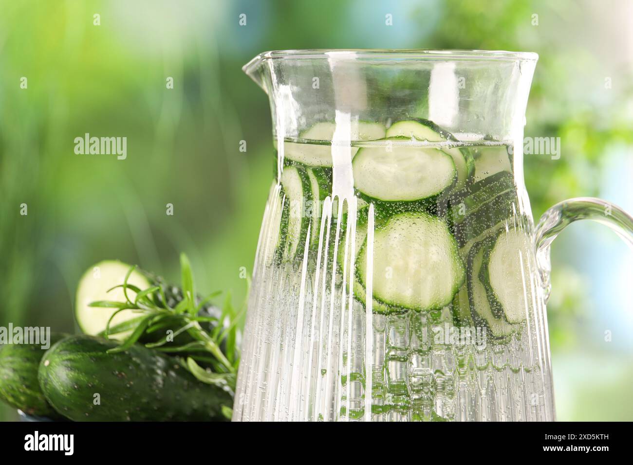 Refreshing cucumber water with rosemary in jug and vegetable against blurred green background ...