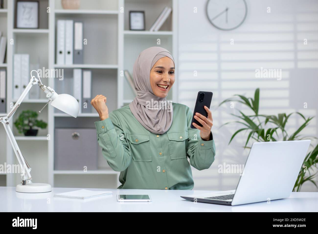 Happy woman in hijab celebrating success while looking at smartphone in ...