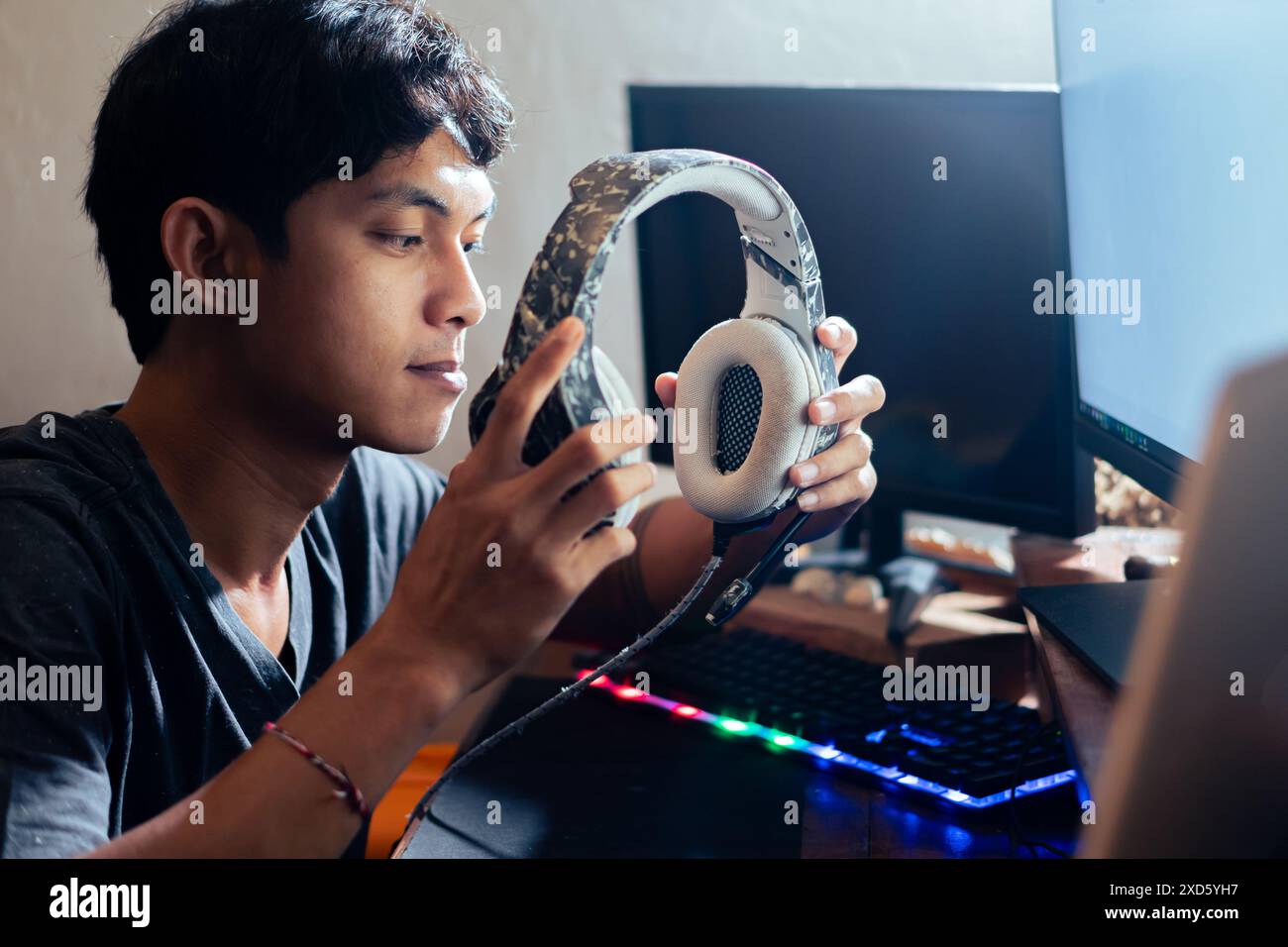 Young man holding gaming headphones in front of a computer setup with RGB keyboard Stock Photo ...