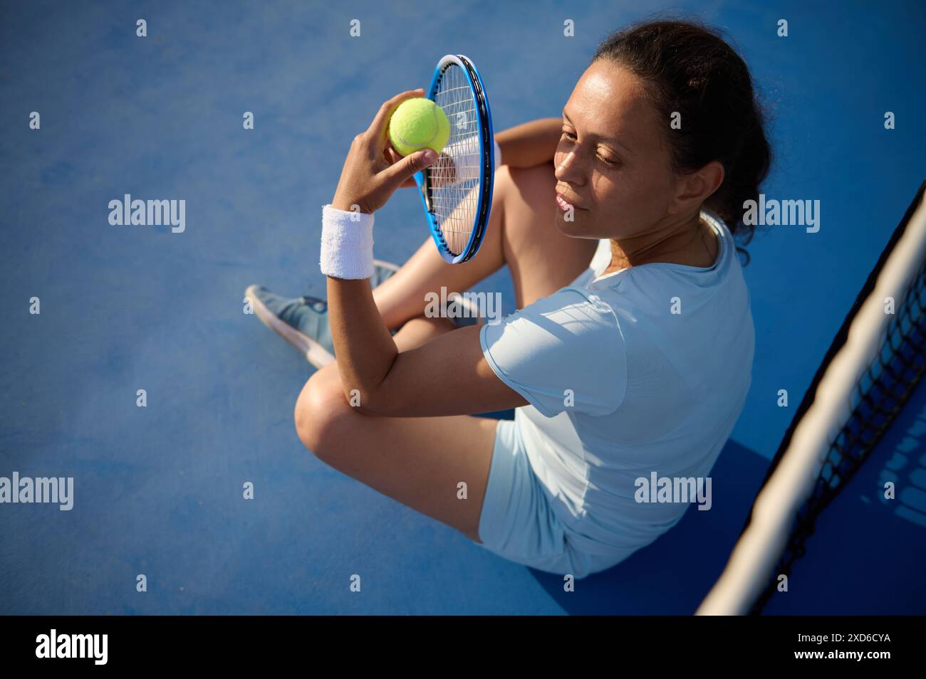 A woman sits on a tennis court holding a tennis racket and ball, taking ...