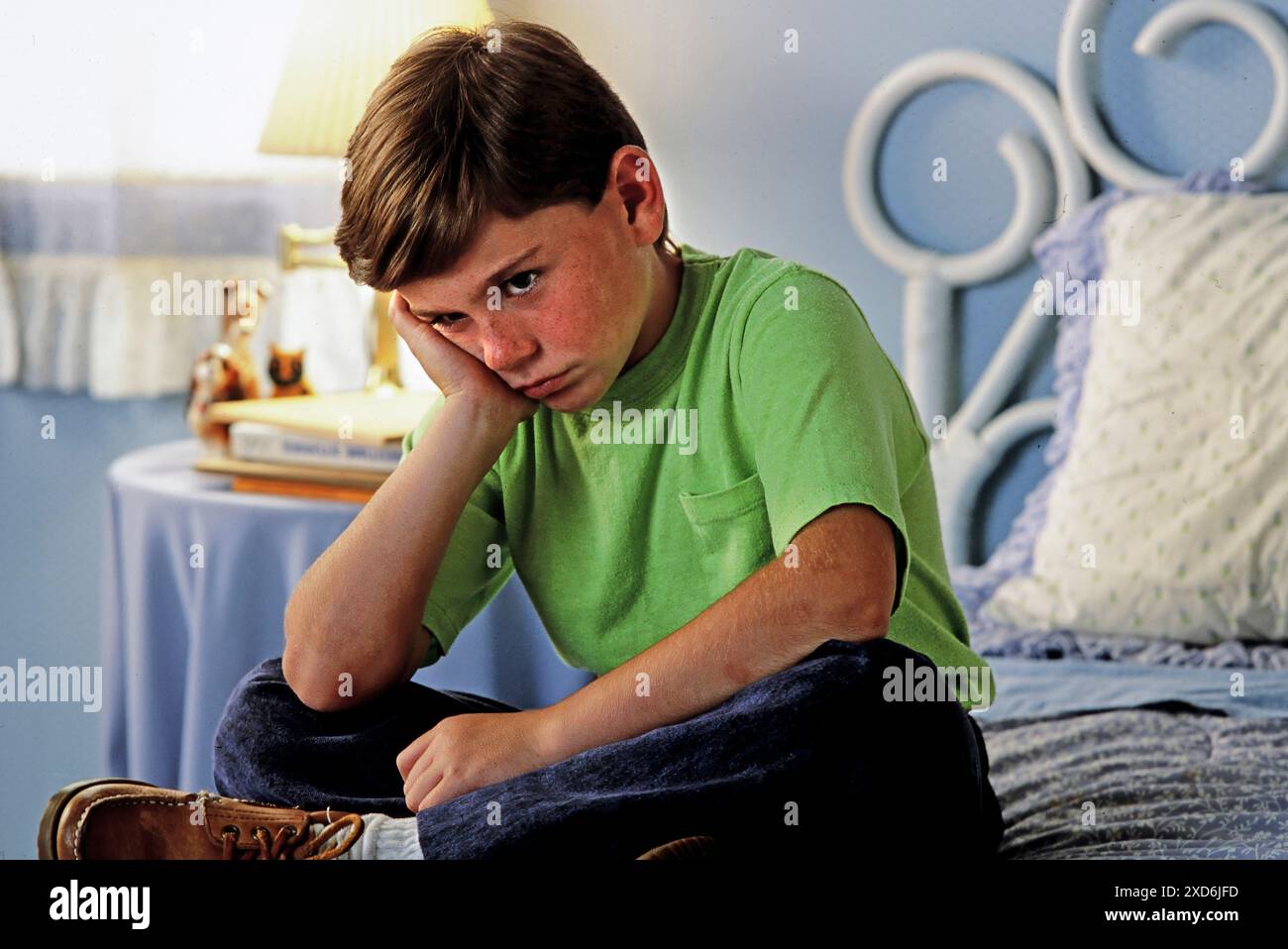 A young boy sits on the edge of a bed with a sad expression on his face ...