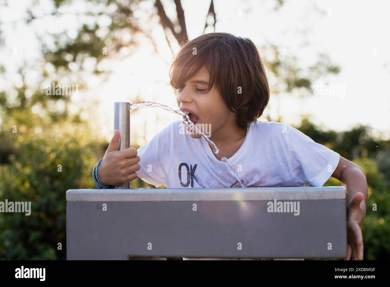 A young boy drinks water from a park fountain on a bright sunny day. He ...