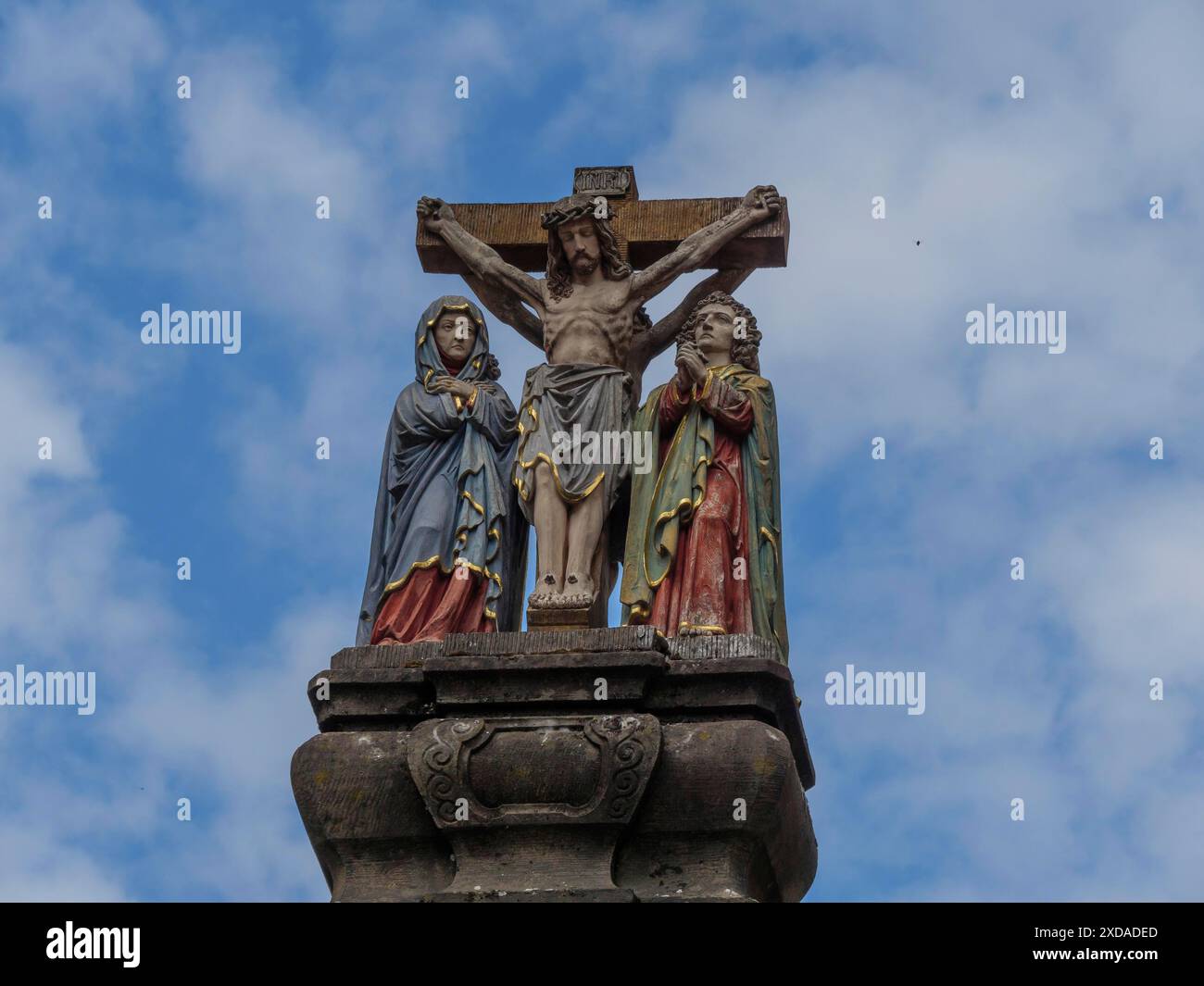 Statue of the crucifixion of Jesus with Mary and another saint below in front of a blue sky ...