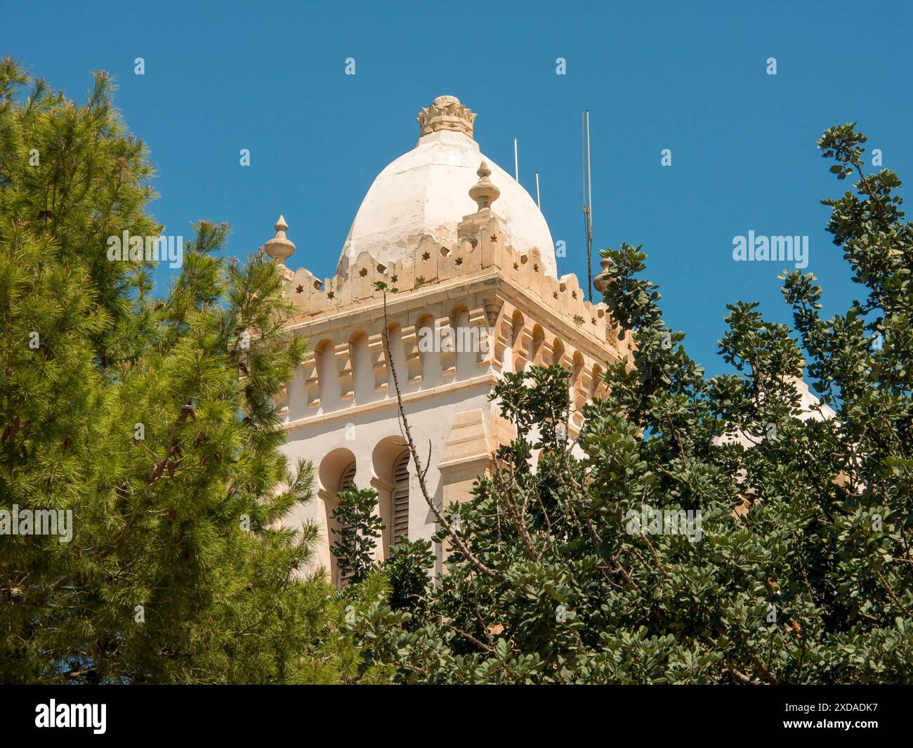 A tower with classical architecture behind dense green trees and a blue ...
