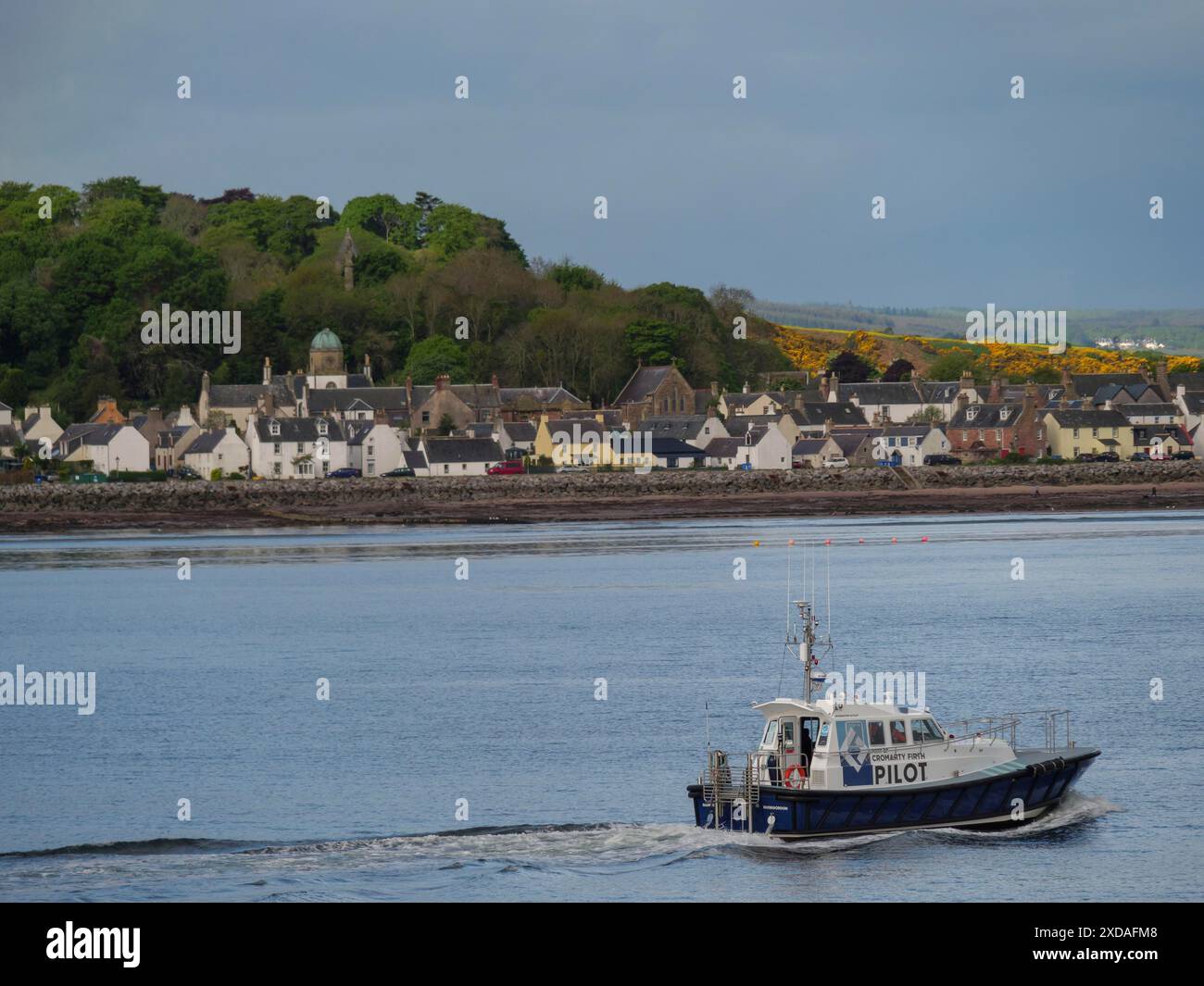 A pilot boat sails on the water while a quiet village of houses lies on ...