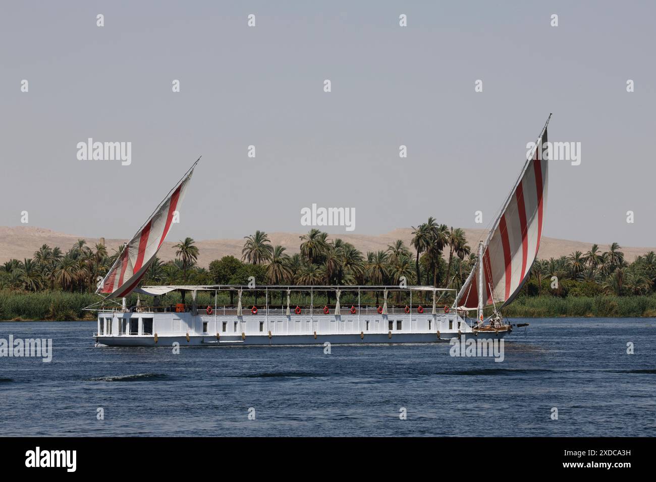 A dahabiya two masted latin-rig sailing boat with red/white striped sails cruises quietly down ...