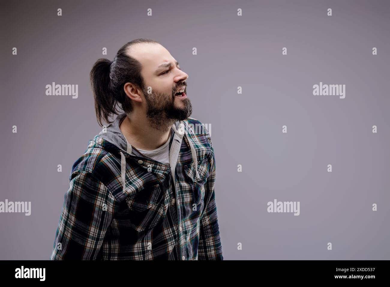 Young man with a beard and ponytail grimaces in pain, looking up and to ...