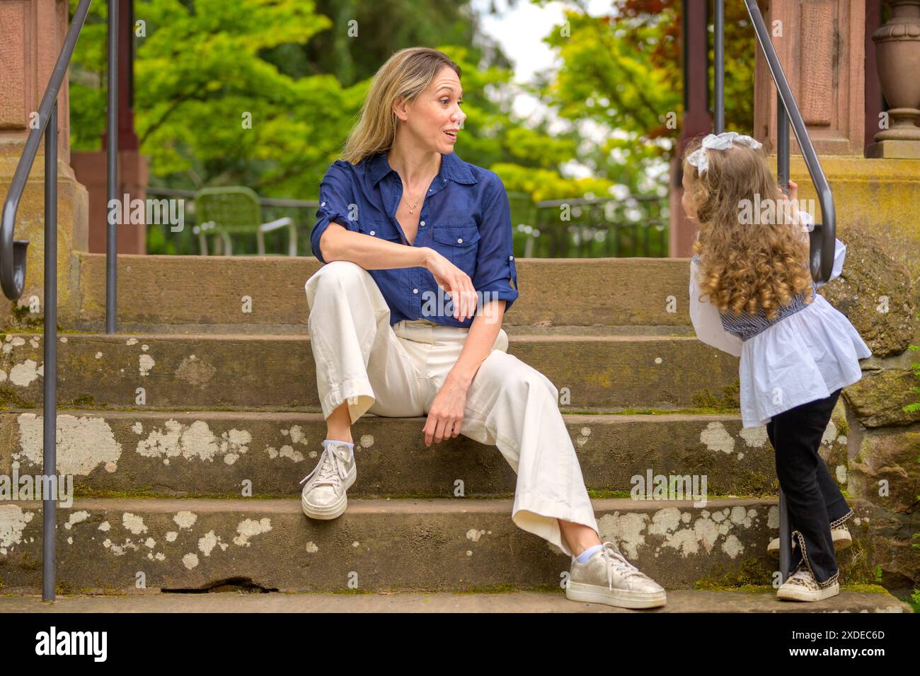 A mother in a blue shirt and white pants sits on outdoor steps ...