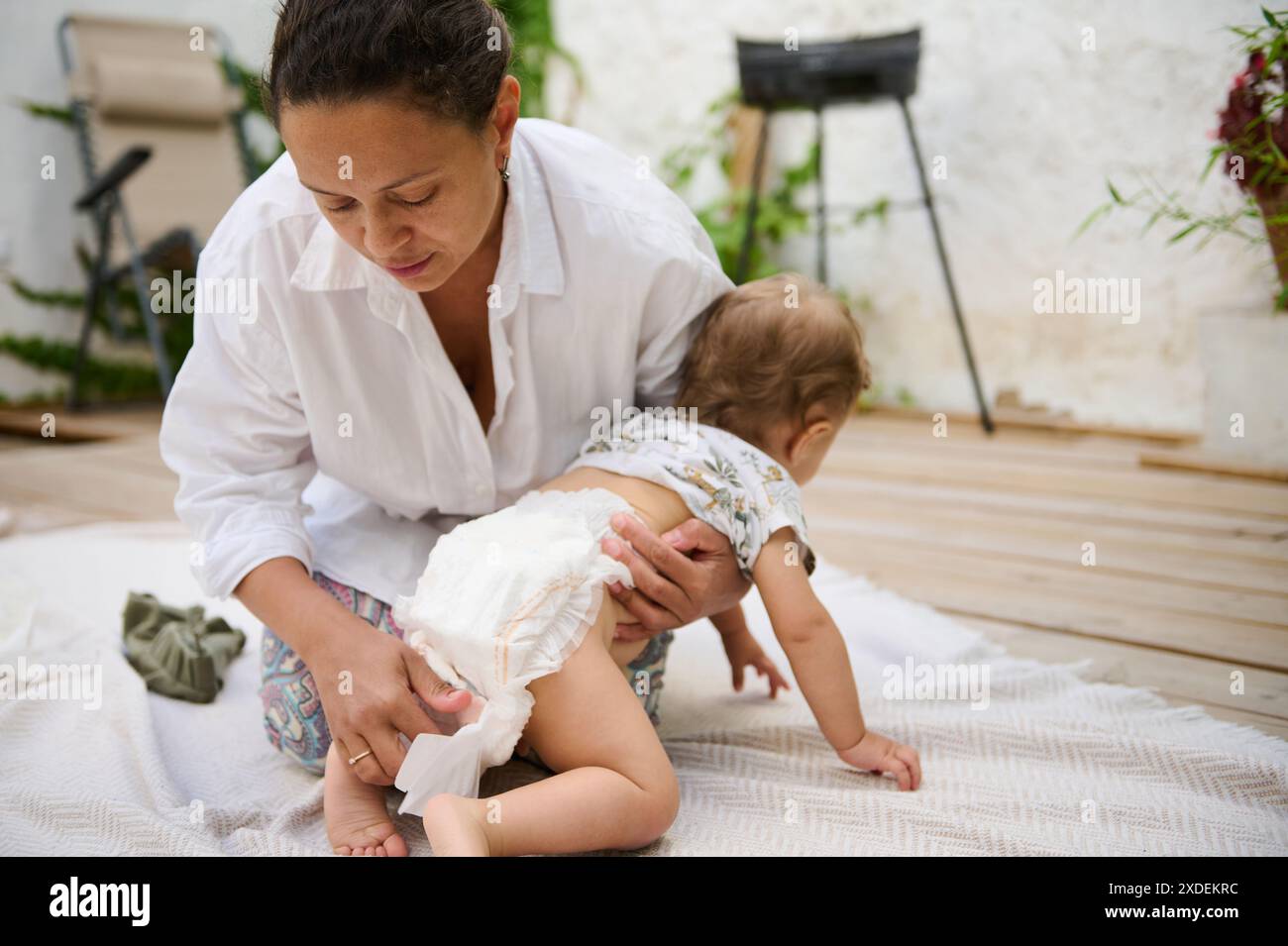 A mother carefully changes her baby's diaper on an outdoor patio ...