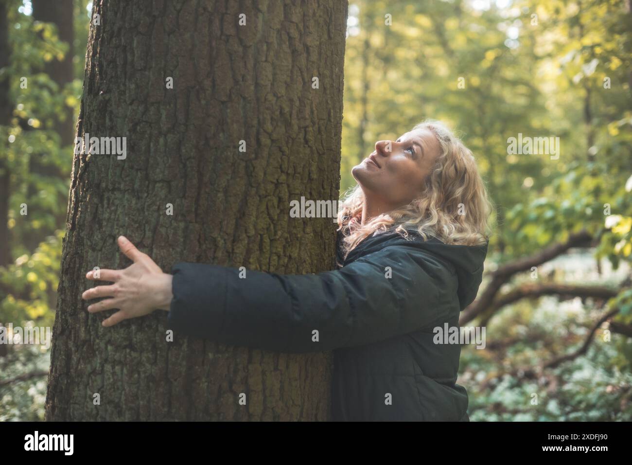 Blonde woman in a long black coat smiles and hugs a massive tree in the ...