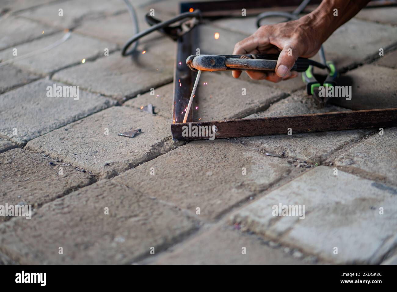 Close-up of a person welding metal rods on a paved surface, with ...
