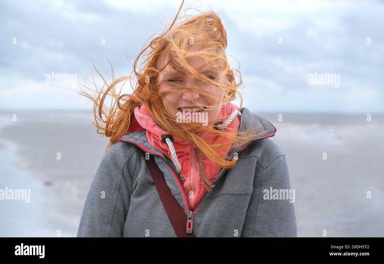 A woman with red hair stands on a windy beach, her hair blowing wildly ...