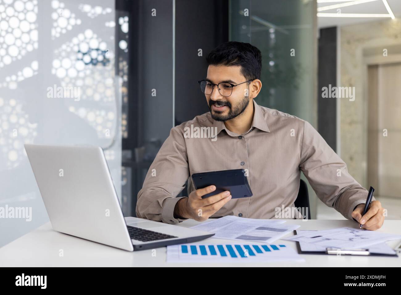 A confident financier uses a calculator and a laptop in a modern office ...